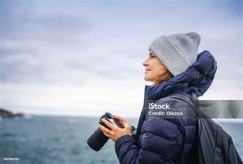 스카프 재킷과 모자를 쓴 젊은 쾌활한 여성 겨울 바다 기슭에 카메라를 손에 들고 여행자 전문 사진 작가 겨울에 대한 스톡 사진