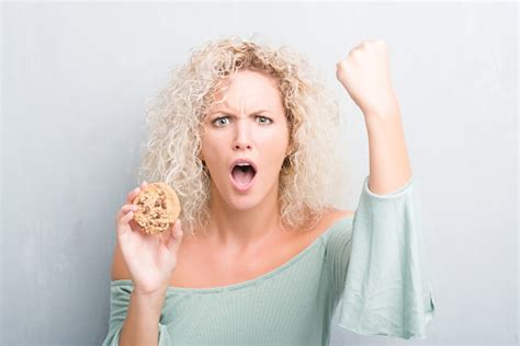 그런 지 회색에 젊은 금발의 여 자가 벽 화가 초콜릿 칩 쿠키 먹고 미친과 제기 손으로 소리 분노와 좌절 외치고 분노 개념
