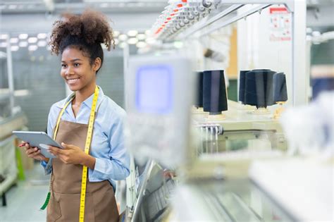 여성 디자이너 측면도 재봉사가 되십시오 직조 산업에서 줄자와 실의 스풀이 있는 패션 디자인 옷을 위한 자신감 있는 미소 짓는 타블렛 작동하는 재봉틀이 있습니다 공장 산업