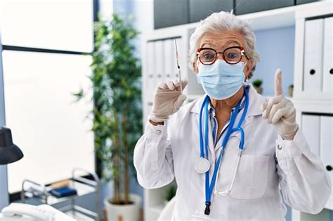의사 유니폼을 입고 회색 머리를 가진 노인 여성과 주사기를 들고 있는 의료 마스크는 행복한 얼굴로 손가락을 가리키는 아이디어나 질문에 놀랐습니다 가리키기에 대한 스톡 사진