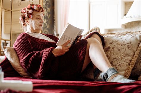 얼굴 마스크와 헤어 컬러 및 독서 책이있는 거실의 소파에 앉아있는 가정 옷을 입은 성숙한 붉은 머리 여성 연금 수령자 가정 레저 및 미용 치료 노인 여성 스킨 케어 노인에