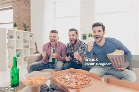 현대 헤어스타일 팝 옥수수 데와 쾌활 한 재미 있는 매력적인 남자의 피자 목표 함 축구 경쟁을 보고 거실에 있는 소파에 앉아 테이블에 칩 3 명에 대한 스톡 사진 및 기타