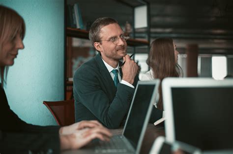 넥타이와 안경을 쓴 양복을 입은 남자가 식탁의 사무실에서 일합니다 동료에 둘러싸여 있습니다 근처에는 노트북을 갖춘 다른 사무실 근로자가 있습니다 고품질 사진 경영자에 대한