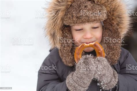 빵과 귀마개가 달린 모자를 쓴 아름다운 작은 러시아 소년의 초상화 한 소년이 공원에서 겨울에 베이글을 먹는다 행복한 어린 시절과