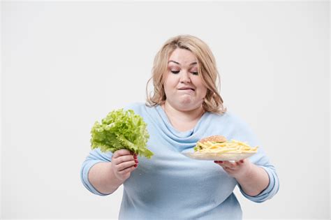 녹색 샐러드 잎과 패스트 푸드 햄버거와 감자 튀김의 접시를 들고 흰색 바탕에 캐주얼 블루 옷에 젊은 무성 한 뚱뚱한 여자