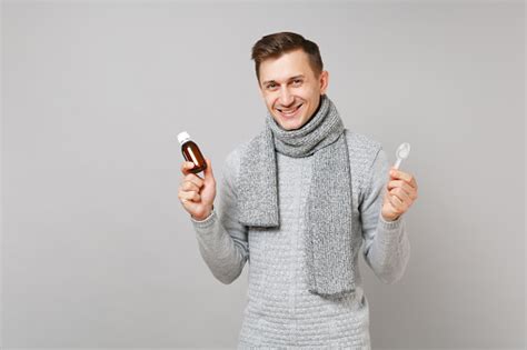 회색 스웨터에 웃는 젊은 남자 스카프 홀드는 병에 액체 약을 붓고 숟가락 회색 배경에 고립 건강 한 라이프 스타일 아픈 질병