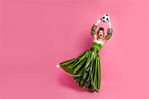 바로크 양식의 녹색 가운을 입은 우아한 여성이 과거와 현대 생활 비교를 보여주는 분홍색 배경에 생생한 표정으로 축구 공을 들고 있습니다 20 24세에 대한 스톡 사진 및