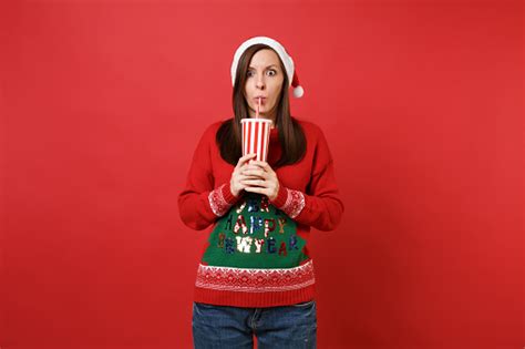 니트 스웨터 크리스마스 모자 마시는 콜라 나 소 다 빨간 배경에 절연 플라스틱 컵에서 산타 소녀 충격 행복 한 새 해 2019 축 하 휴일 파티 개념입니다 복사 공간을