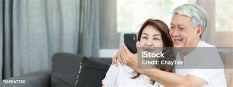 매력적인 오래된 아시아 커플 남편과 아내의 솔직한 우리 기술 스마트 폰 5g 인터넷 연결을 통해 집에서 코치에 앉아 선임 부모가 모바일 화면을 보거나 보고 있습니다 커플에