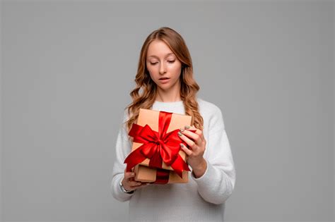 깜짝 놀란 표정으로 선물 상자 안을 들여다보는 흰색 스웨터를 입은 젊은 여성은 비밀리에 선물을 풀고 회색 배경 위에 서 있었다 새해 여성의 날 생일 휴일 컨셉 25 29세에