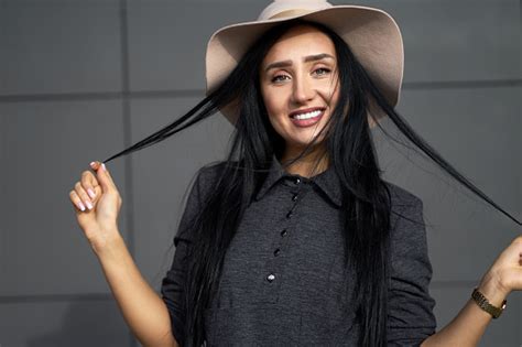 봄 모던 컬렉션 세련된 회색 드레스와 세련된 모자를 입고 행복 아름다운 갈색 머리 소녀 회색 도시 벽 배경에 포즈를 취하는 트렌디 한 의상을 입은 모델 야외 초상화입니다