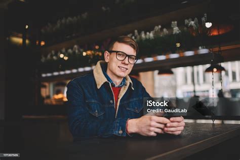 세련된 커피숍에서 휴식을 취하며 스마트폰으로 소셜 네트워크에서 프로필을 업데이트하는 동안 카메라에 미소 짓는 데님 재킷을 입은 성공적인 남성 블로거의 절반 길이 초상화 4g