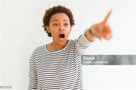 젊은 아름 다운 아프리카계 미국인 여자 흰색 배경 위에 줄무늬 스웨터를 입고 앞에 놀란 손가락으로 오픈 입 깜짝 표현 앞에 뭔가 가리키기에 대한 스톡 사진 및 기타 이미지