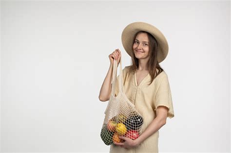 가벼운 드레스를 입고 챙이 넓은 밀짚모자를 쓴 여성이 형형색색의 과일이 가득 담긴 그물 가방을 들고 미소를 짓고 있다 그녀는 평범한 배경에 서 있다 30 34세에 대한 스톡
