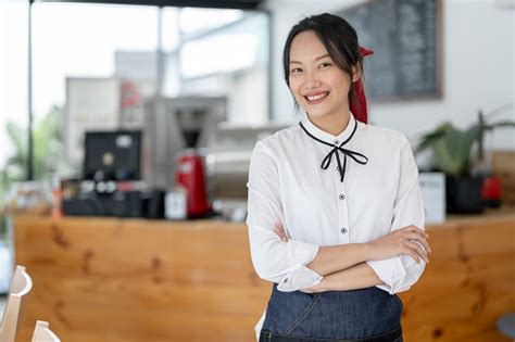 커피 비즈니스 소유자 앞치마에있는 매력적인 젊은 아름다운 백인 바리스타와 커피 숍 카운터의 카메라에 웃고있는 십자가 손 경영자에