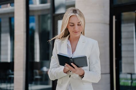 흰 양복을 입은 금발의 백인 사업가가 노트를 들고 미소를 지으며 독서 의제를 읽고 거리에 서서 회의에 참석합니다 금융 보조원은 작업 목록을 확인하고 클라이언트를 기다리고