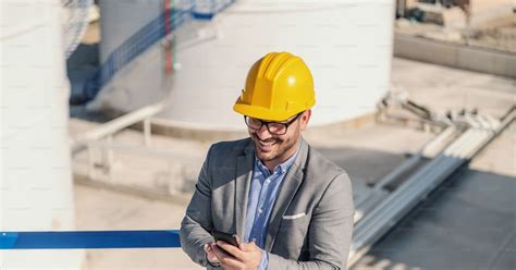 난간에 기대어 스마트 폰을 사용하는 머리에 헬멧을 쓴 양복을 입은 성공적인 매력적인 우아한 부유 한 사업가를 웃고 있습니다 정유소 외관 사진 Unsplash의 석유