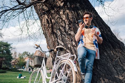 스마트 폰을 사용하고 자전거와 함께 공원의 나무 아래에서 커피를 마시는 젊은이 20 29세에 대한 스톡 사진 및 기타 이미지 20 29세 30 34세 Brand