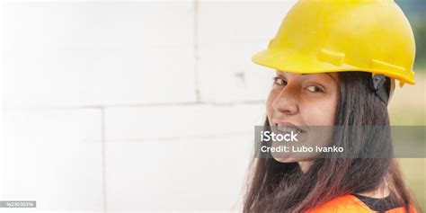 노란색 안전모와 주황색 눈에 잘 띄는 조끼를 입은 젊은 여성 긴 검은 머리 어깨 너머로 바라보며 자신 있게 웃고 있다 흐릿한 건설 현장 벽 텍스트 왼쪽 공간 25 29세에
