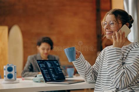 Young Female Computer Programmer Is Talking Phone While Coding At Laptop Sitting In Coworking