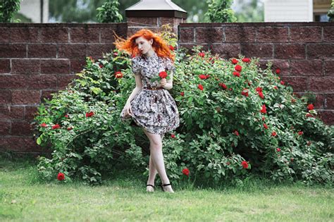 빨간 장미와 부시 대통령의 배경에서 꽃 드레스에 붉은 곱슬 머리를 가진 여자 창백한 피부 파란 눈 밝은 특이 한 모양을 하 고 붉은 입술과 정원에서 얇은 허리와 빨간 머리