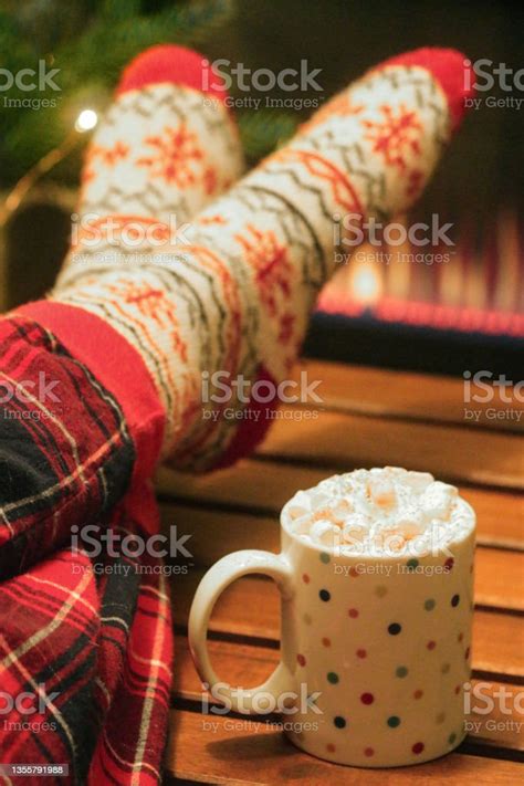 빨간 타탄 잠옷과 크리스마스 테마 패턴 양말 커피 테이블에 발 발목 얼룩 핫 초콜릿 찻잔 휘핑 크림과 미니 마시멜로를 얹은 가스 불꽃의 행을 입고 인식 할 수없는 여성의