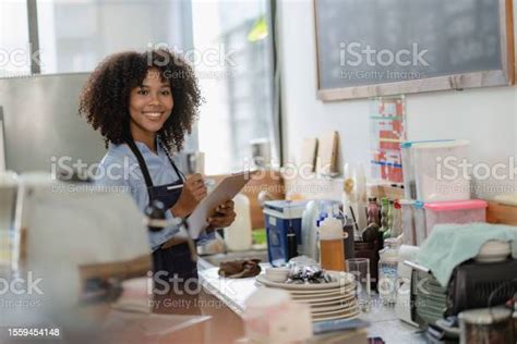 아름다운 아프리카계 미국인 여성 계산원 또는 바리스타가 구매 세부 정보를 작성하고 가용성을 확인합니다 소상공인 앞치마를 입은 커피숍이 카운터 앞에 서 있다 20 29세에