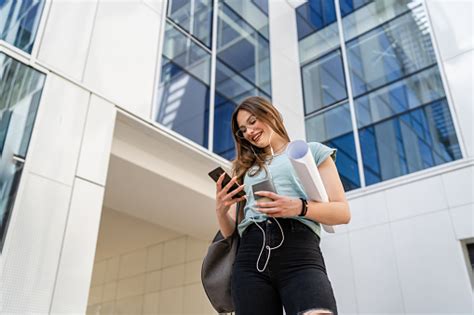 한 여성 젊은 성인 백인 여학생이 종이와 커피 한 잔을 들고 대학 앞에 서서 행복한 미소를 기다리는 휴대 전화를 사용하여 야외에서 행복한 미소를 짓고 실제 사람들이 공간을