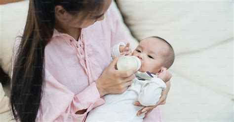 아시아의 젊은 어머니는 침실에 있는 우유병에서 귀여운 신생아를 안고 먹이를 줍니다 소녀는 따뜻한 미소로 귀여운 소년을 돌 봅니다 행복한 소년은 사랑스러운 집에서 엄마와