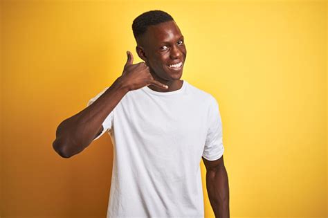 고립 된 노란색 배경 위에 서있는 흰색 티셔츠를 입은 젊은 아프리카 계 미국인 남성이 전화 통화처럼 손과 손가락으로 전화 제스처를하고 미소 짓고 있습니다 개념을 전달합니다