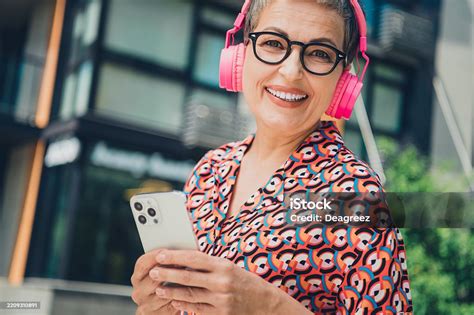 도시 공원에서 화창한 날 야외에서 분홍색 헤드폰과 스마트 폰으로 음악을 즐기는 노인 여성 50 54세에 대한 스톡 사진 및 기타 이미지 50 54세 Brand Name