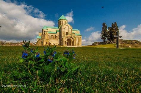 A Large Building With A Green Roof And Blue Flowers In The Foreground On A Sunny Day