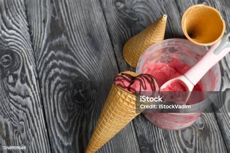와플 콘에 딸기 아이스크림 초콜릿으로 장식 근처에는 아이스크림이 없는 빈 와플 콘이 있습니다 아이스크림 용기와 숟가락 검은 색과 흰색으로 그려진 소나무 보드에 냉동된에 대한