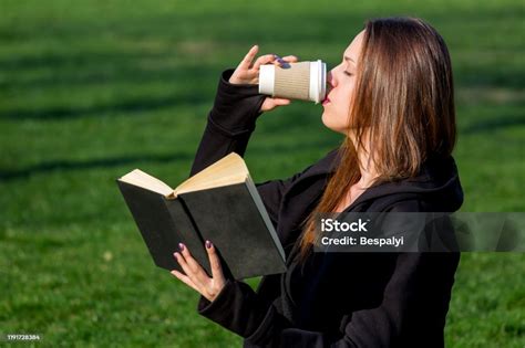 검은 코트와 검은 색 하드 커버 책에 긴 머리를 가진 갈색 머리 소녀의 초상화는 손에 녹색 잔디밭에 화창한 날에 일회용 종이 스택에서 뜨거운 커피를 마십니다 갈색 머리에