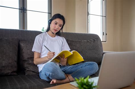 아시아 행복 한 젊은 여자 학생 착용 헤드폰 공부 온라인 보고 Webinar 팟 캐스트 에 노트북 듣기 교육 과정 컨퍼런스 전화 및 쓰기 노트 집에서 소파에 앉아 노트북에
