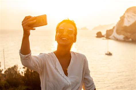 폰자 섬 해안의 바다 앞에서 일몰과 함께 셀카를 찍고있는 스마트 폰을 가진 젊은 여성이 바다를 볼 수있는 벽에 있습니다 Date Night Romance에 대한 스톡