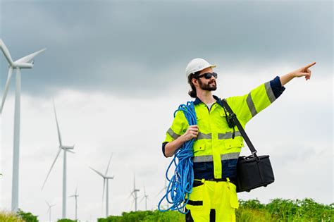 밝은 안전 장비를 착용한 작업자가 재생 에너지 프로젝트 중 흐린 하늘 아래 풍력 터빈을 가리키고 있습니다 건설 산업에 대한 스톡 사진 및 기타 이미지 Istock