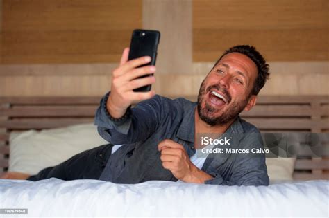 젊은 매력적이 고 행복 한 30 대 또는 40 대 남자 촬영 Selfie 사진 휴대 전화 웃는 장난 몸짓에서 홈 침실 침대에 누워 캐주얼에 소셜 미디어 재미의 라이프 스타일