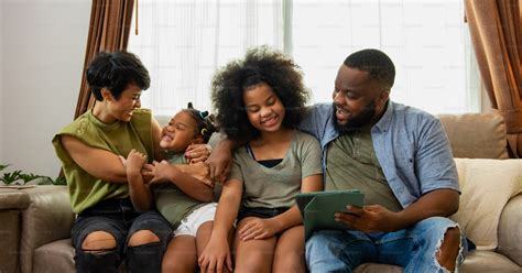 아프리카 혼혈 부모와 두 어린 딸이 거실 소파에 앉아 디지털 태블릿을 사용하여 게임을 하거나 함께 영화를 봅니다 행복한 가족은 집에서 함께 기술로 주말 활동을 즐깁니다