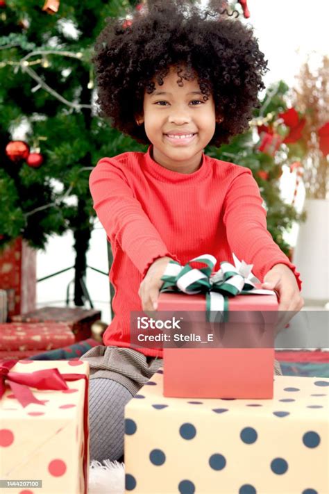 장식 크리스마스 트리 아래에서 크리스마스 선물 선물 상자를 들고 검은 카레 머리를 가진 사랑스러운 행복한 미소를 짓는 아프리카계 미국인 소녀 아이의 초상화 행복한 크리스마스
