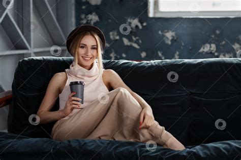 Модная красивая молодая девушка сидит на диване со стаканом свежего кофе в уютной атмосфере