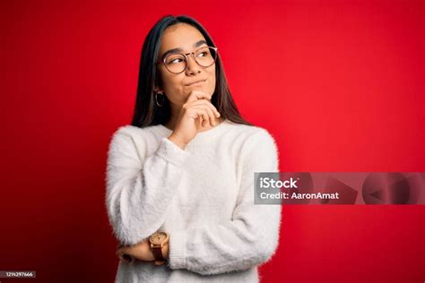 젊은 아름 다운 아시아 여자 캐주얼 스웨터와 안경을 입고 빨간 배경 에 턱에 대 한 생각 펜시브 표정 사려 깊은 얼굴로 미소 의심 개념 갈색 머리에 대한 스톡 사진 및 기타