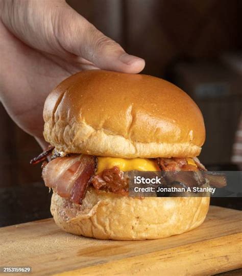나무 테이블 위에 브리오슈 빵 고기 체다 치즈 바비큐 소스 베이컨 조각이 놓인 커다란 햄버거를 손에 뻗는 손 건강하지 못한 생활방식에 대한 스톡 사진 및 기타 이미지