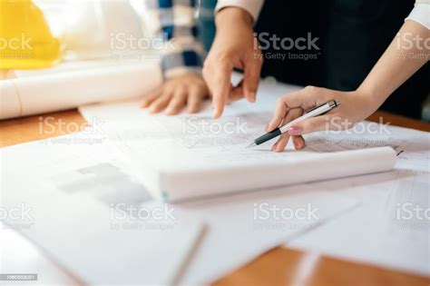 건축가 또는 엔지니어는 아이디어를 브레인스토밍하는 데 도움을 주고 구조 설계 및 모델 시공을 위해 파트너와 함께 만나 작업장의 테이블에 있는 청사진을 가리킵니다 건설 현장에