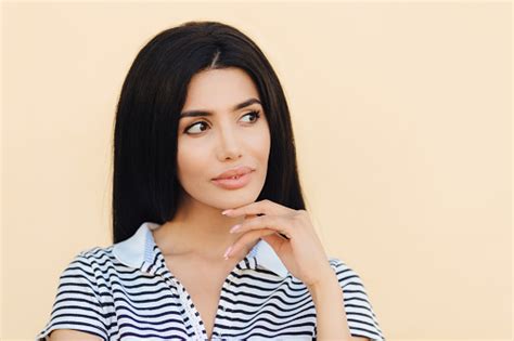 건강한 피부를 가진 매력적인 갈색 머리 여성의 초상화를 닫고 옆으로 가볍게 보이고 턱 아래에 손을 대고 밝은 분홍색 배경 위에 고립 된 즐거운 무언가에 대한 꿈을 꾸십시오