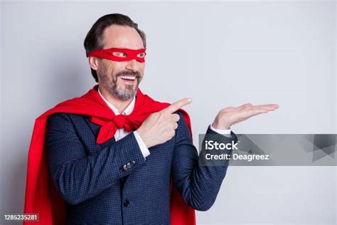 사진 쾌활한 성숙한 사업가 슈퍼 히어로 의상 직접 손가락 오픈 팔 홀드 참신한 제품 빈 공간 제공 정장 빨간 얼굴 마스크 망토 고립 된 회색 배경 남자에 대한 스톡 사진 및