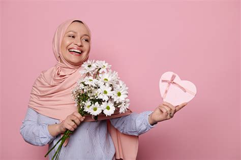 히잡을 쓴 쾌활한 무슬림 여성 꽃다발과 선물 상자를 들고 고립된 분홍색 배경을 옆으로 바라보며 웃고 있다 International Womens Day에 대한 스톡 사진 및