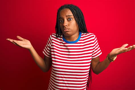 줄무늬 티셔츠를 입은 젊은 아프리카계 미국인 여성이 고립된 빨간 배경 위에 서서 팔과 손을 들고 혼란스러운 표정을 짓고 있다 의심 개념 땋은 머리에 대한 스톡 사진 및 기타