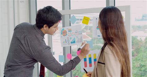 비즈니스 사람들은 사무실의 프로젝트 계획 보드에서 일하고 동료 친구와 대화하여 프로젝트 개발을 분석합니다 그들은 유리 벽에 게시 된 스티커 메모를 사용하여 정리합니다