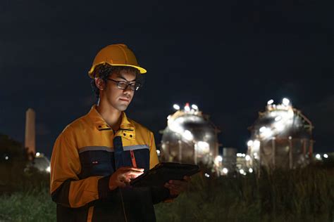 안전모와 노란색 안전 유니폼을 입은 중년 산업 엔지니어가 밤에는 조명이 켜진 정유 산업 공장을 배경으로 디지털 태블릿을 통해 원격으로 생산 과정을 검사하고 있습니다 밤의